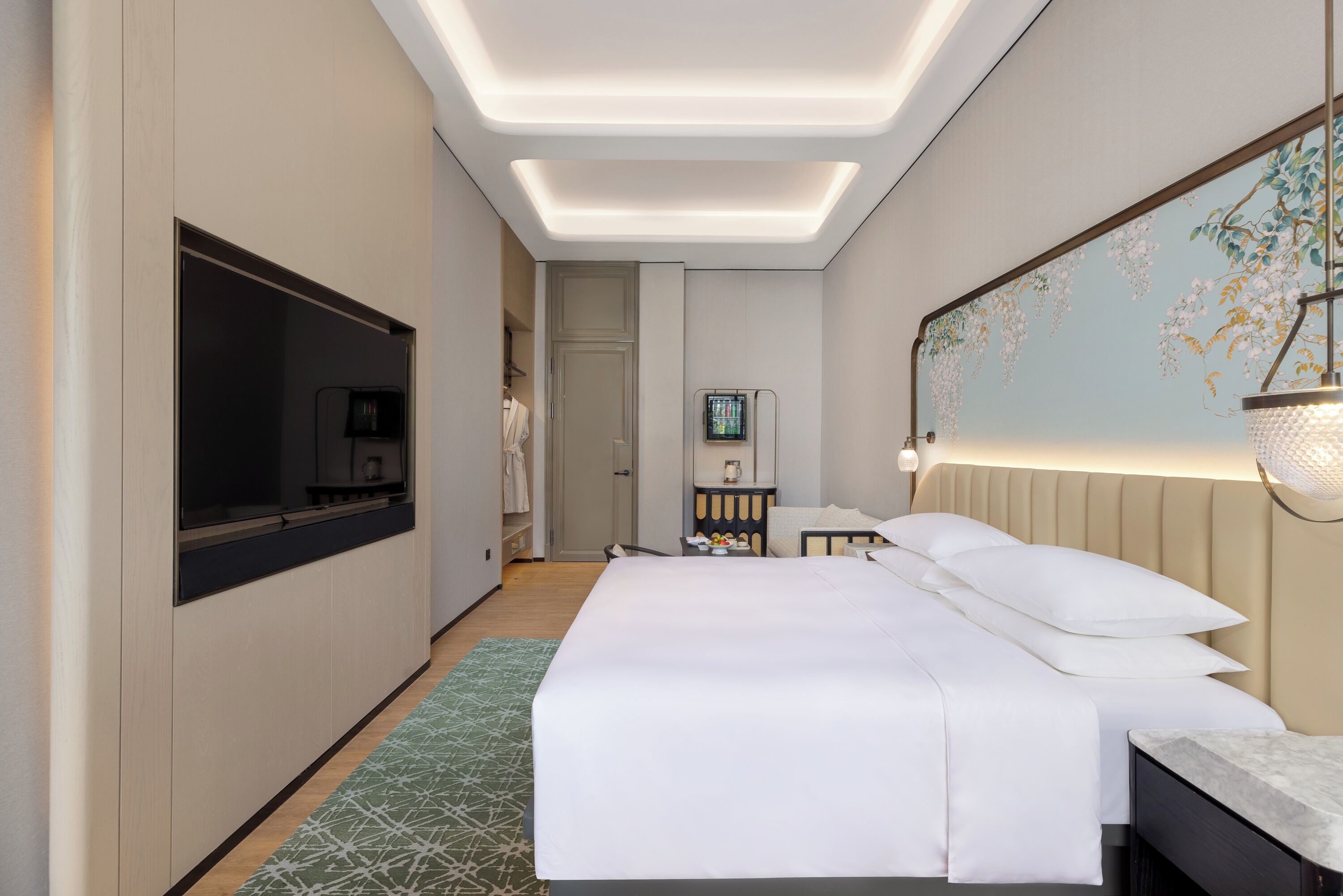 Quarto de hotel moderno com uma cama grande, uma TV de tela plana na parede, uma mesa de cabeceira com abajur e um detalhe decorativo na parede.
