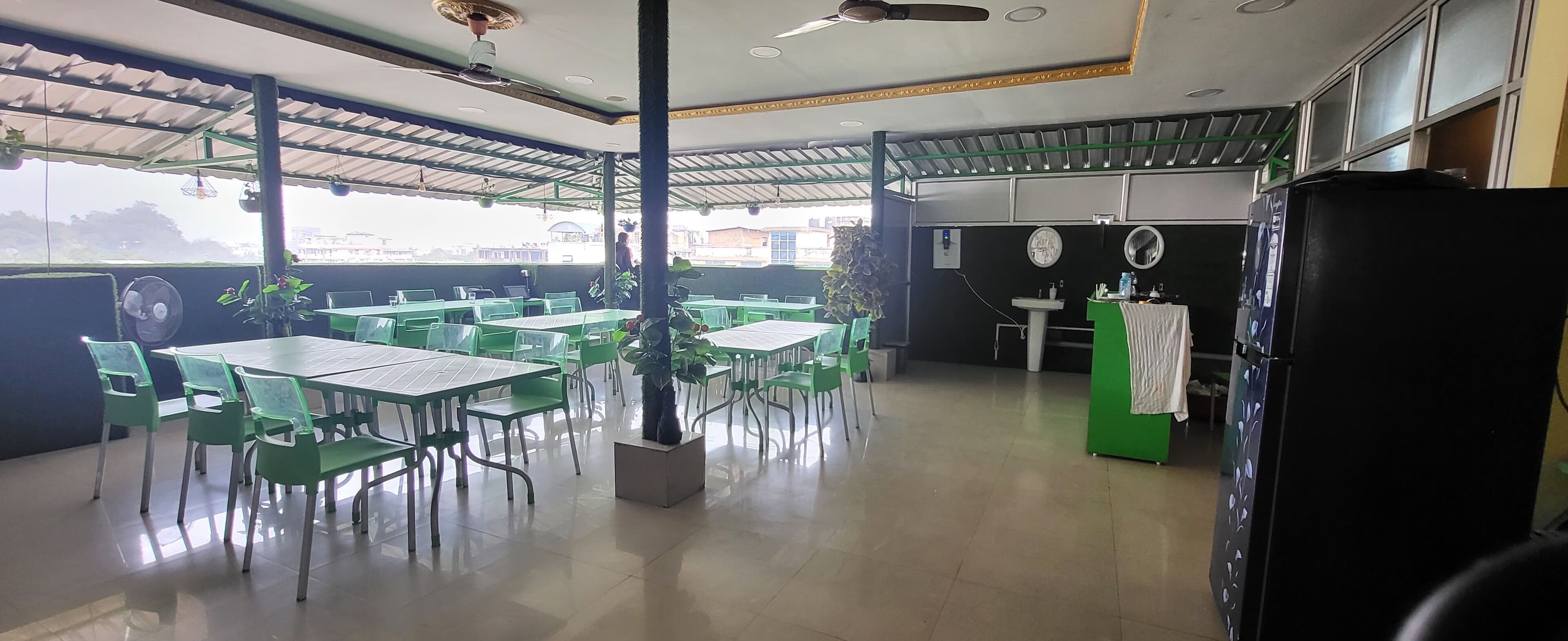 Restaurante com mesas e cadeiras verdes, um ventilador de teto e vista para edifícios lá fora.