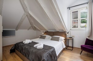 Quarto com teto inclinado, cama com colcha estampada, duas almofadas, mesa de cabeceira com abajur e janela com persianas.
