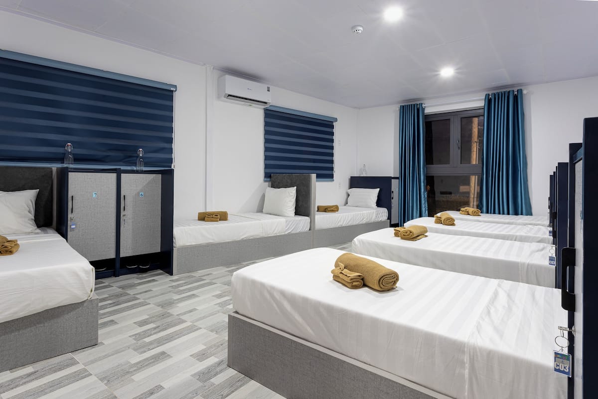 Quarto de hotel com três camas de solteiro, um guarda-roupa, um aparelho de ar condicionado e uma janela com cortinas.