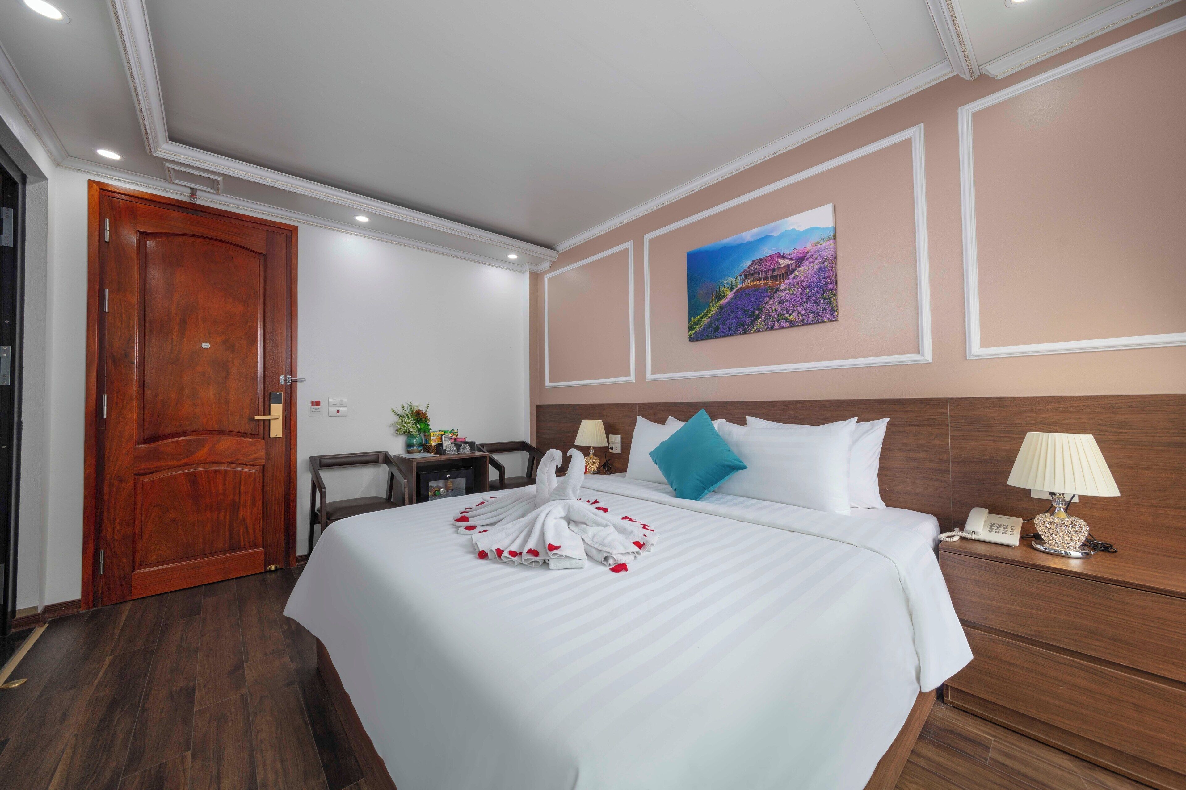 Quarto de hotel com uma cama grande, porta de madeira, mesa de cabeceira com abajur e um quadro na parede.