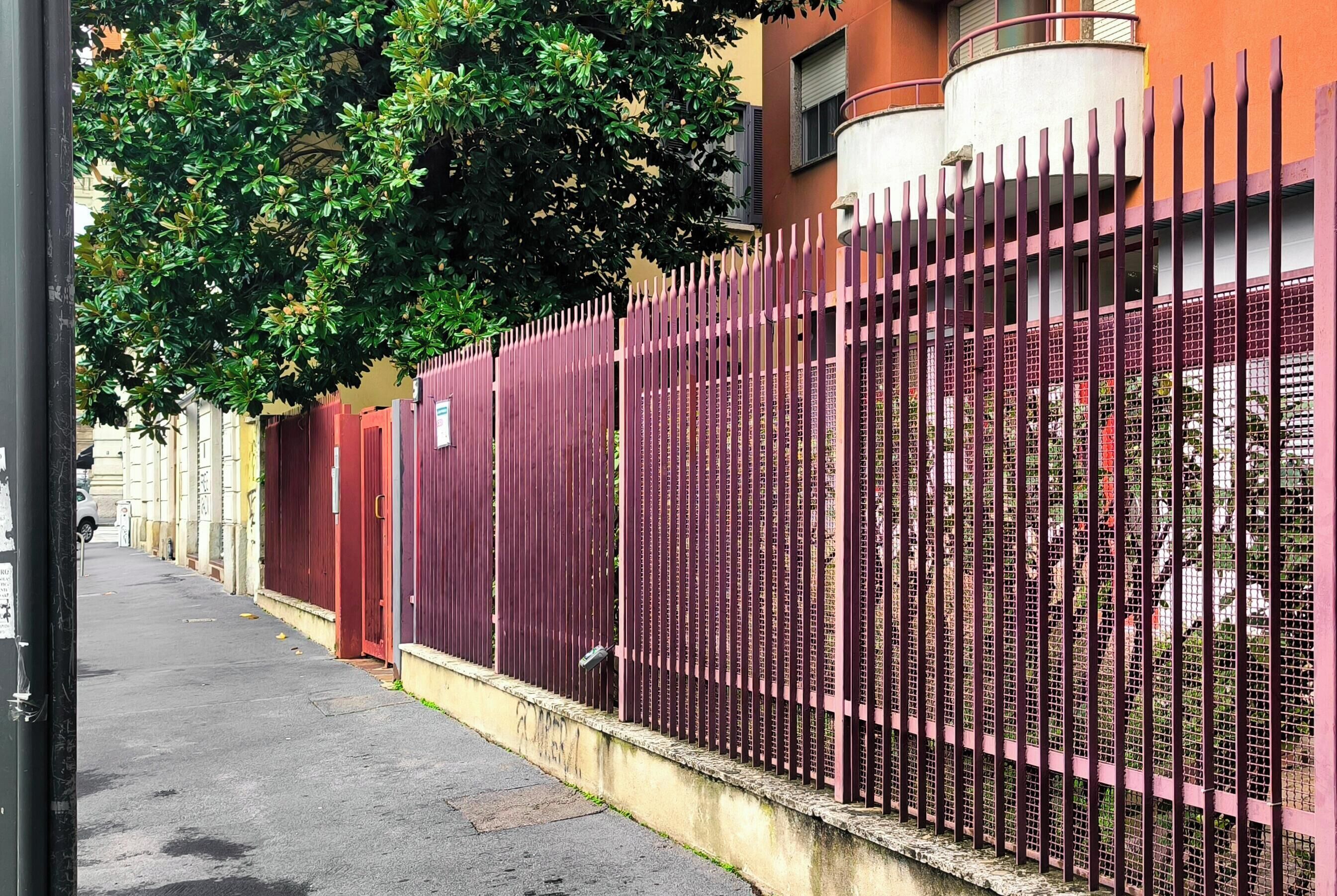 Rua com cerca de metal vermelha, uma árvore e edifícios ao fundo.