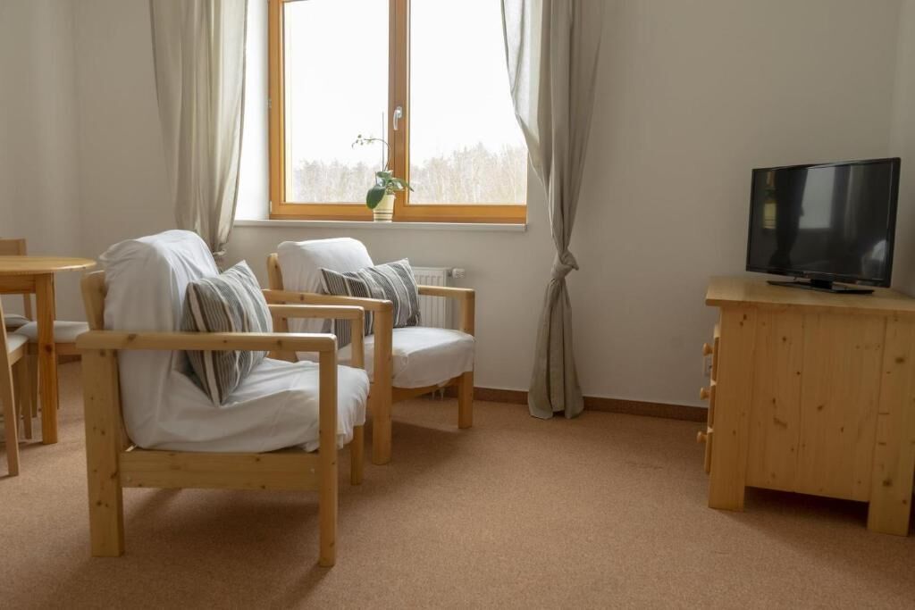 Quarto com duas cadeiras de madeira, uma pequena mesa de madeira, uma televisão sobre um suporte de madeira e uma janela com cortinas.