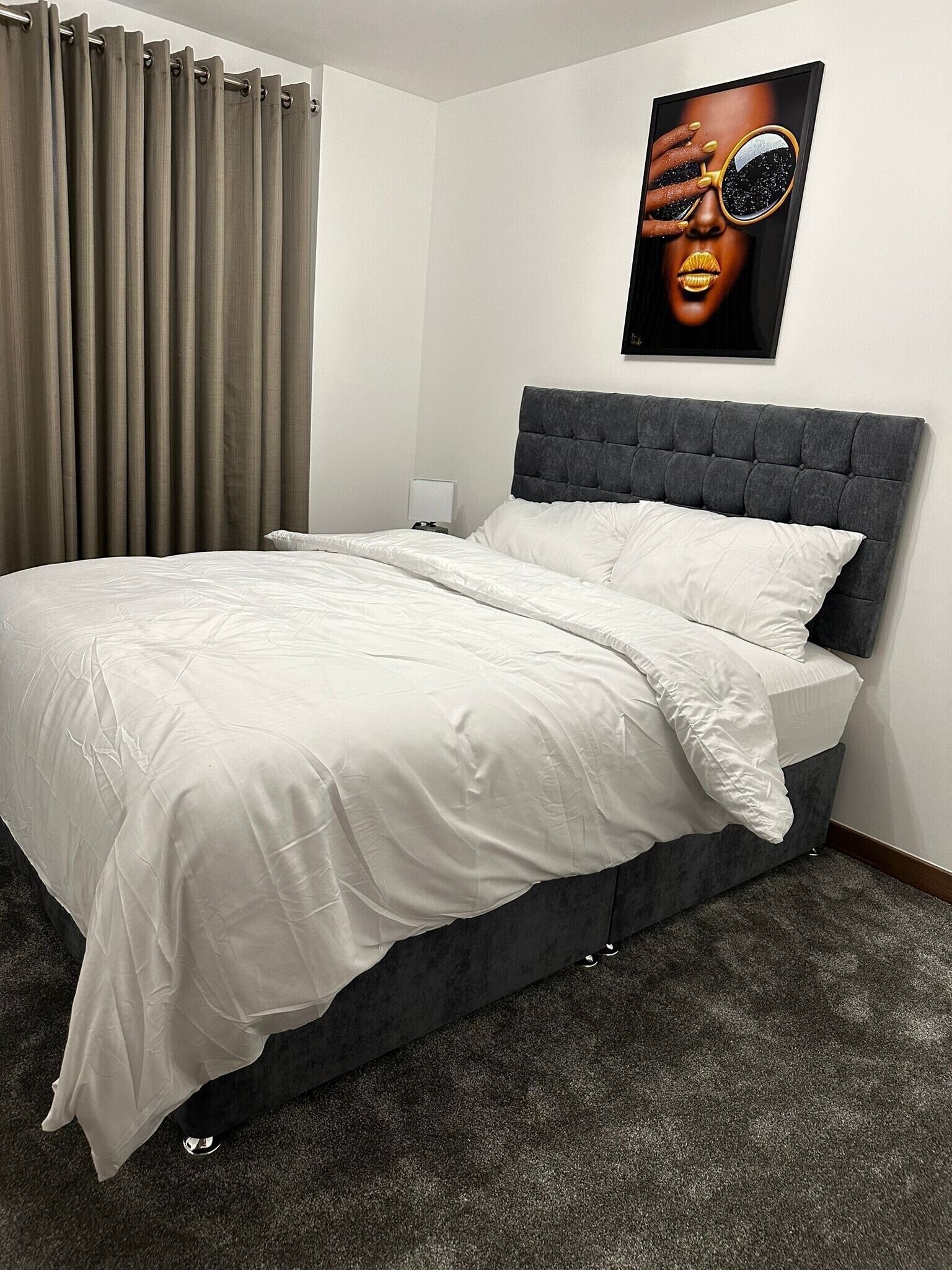 Quarto com cabeceira estofada cinza, roupa de cama branca e quadro pendurado na parede.