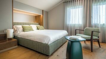 Quarto de hotel moderno com uma cama grande, uma poltrona estofada em verde, uma mesa redonda pequena e piso de madeira.