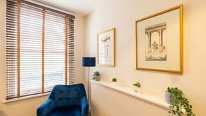 Um quarto com uma cadeira azul, uma janela com persianas, um abajur e duas molduras com quadros na parede.