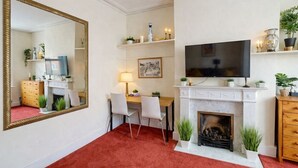 Quarto de hotel com um espelho grande, lareira, televisão, escrivaninha com cadeiras e um tapete vermelho.
