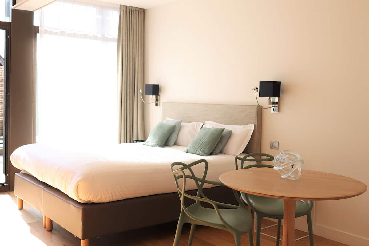 Quarto de hotel moderno com uma cama grande, duas luminárias fixadas na parede, uma mesa redonda de madeira e vista para a cidade.