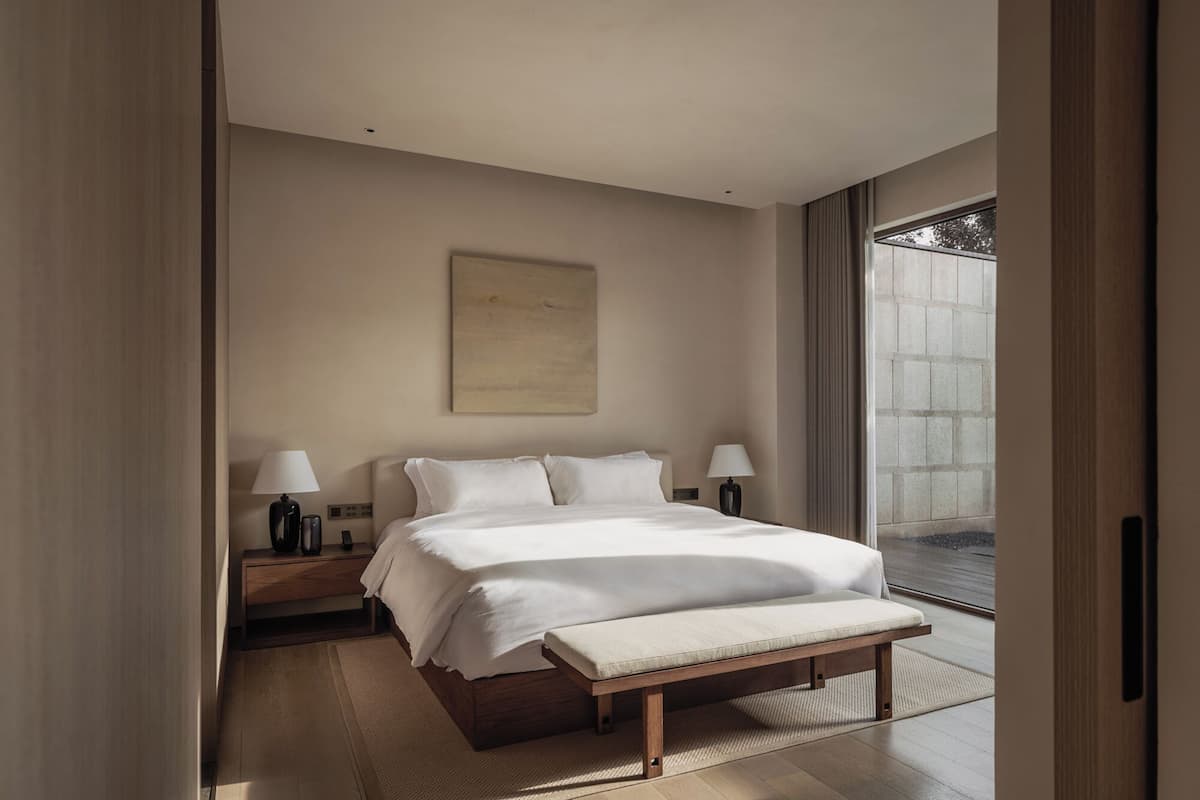 Quarto moderno com uma cama grande, mesas de cabeceira, um banquinho e vista para o exterior.
