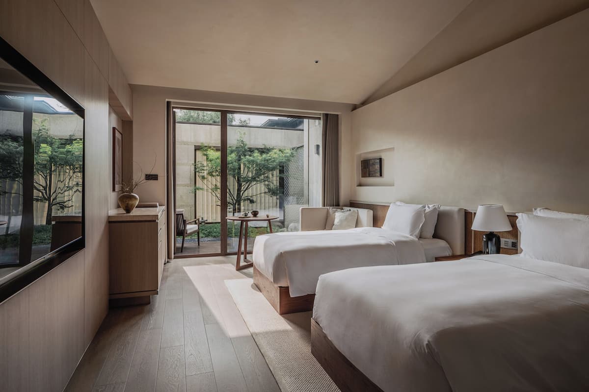 Quarto de hotel moderno com duas camas, uma mesa pequena e vista para um jardim através de janelas amplas.