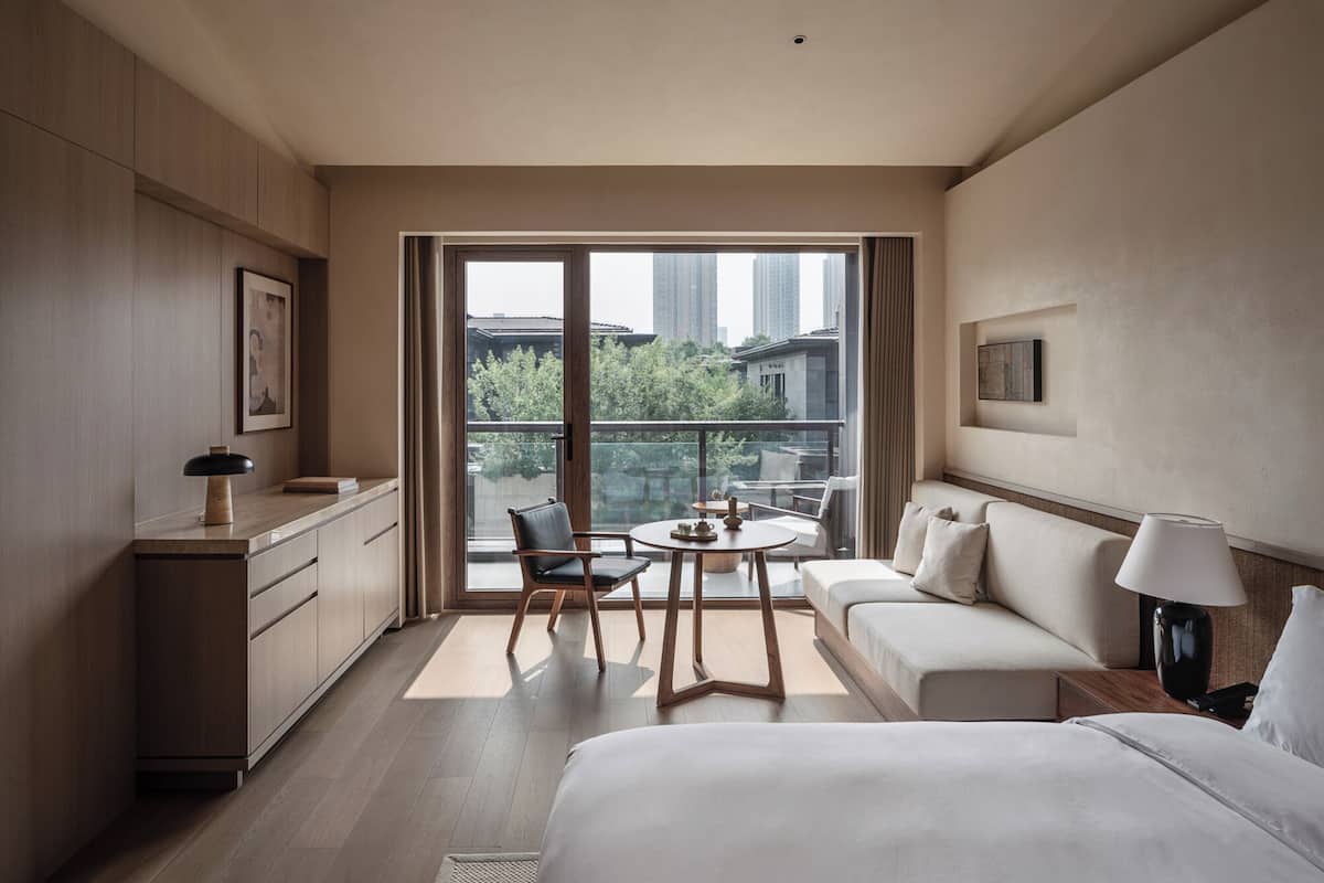 Quarto de hotel moderno com uma cama grande, um sofá, uma mesa de jantar e uma varanda com vista para a cidade.