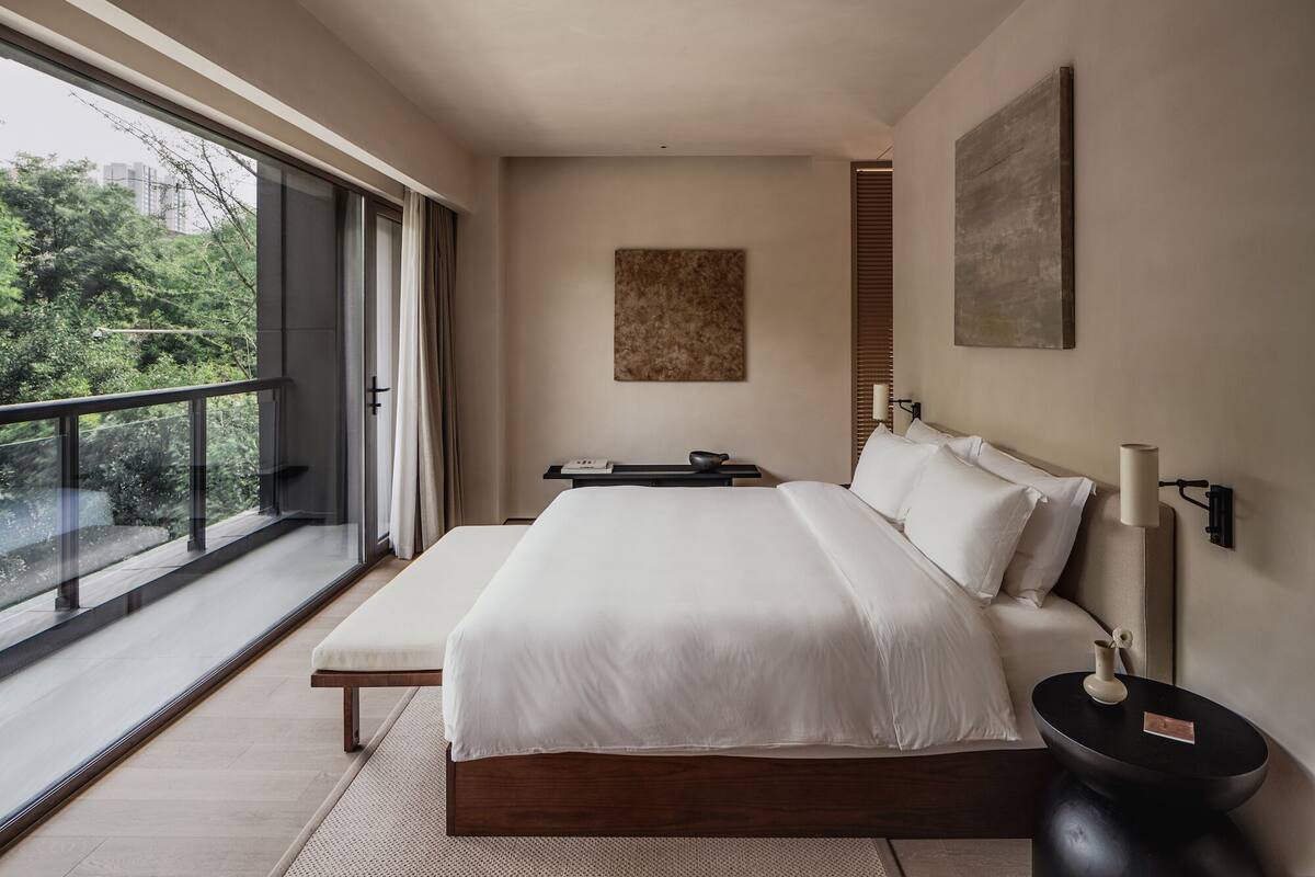 Quarto moderno com uma cama grande, mesa de cabeceira, um banquinho e vista para o verde lá fora.