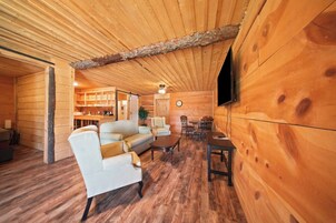Interior de uma cabana de tronco com sala de jantar, sofá, televisão e paredes de madeira.