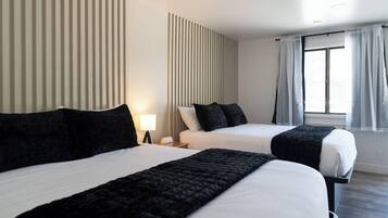 Quarto de hotel com duas camas, uma mesa de cabeceira com abajur, uma janela com cortinas e um painel com persianas verticais.