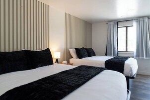Quarto de hotel com duas camas, uma mesa de cabeceira com abajur, uma janela com cortinas e um painel com persianas verticais.