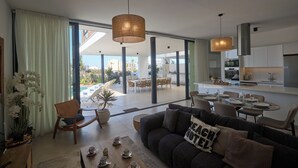 Uma sala de estar moderna com sofá, mesa de jantar e cadeiras, cozinha e vista para uma piscina e área de estar externa.
