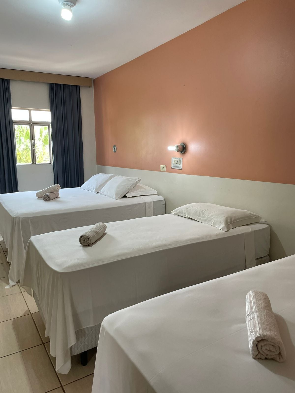 Quarto de hotel com duas camas de solteiro, janela com cortinas, luminária na parede e ventoinha no teto.