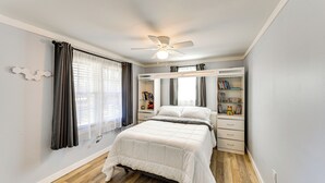 Quarto com cama, ventoinha no teto, janela com cortinas e estantes embutidas com livros e objetos decorativos.