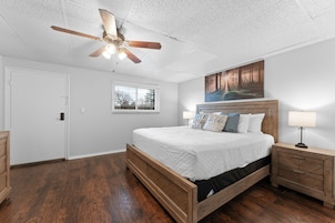 Quarto com cama de madeira, ventilador de teto, janela e um quadro na parede.