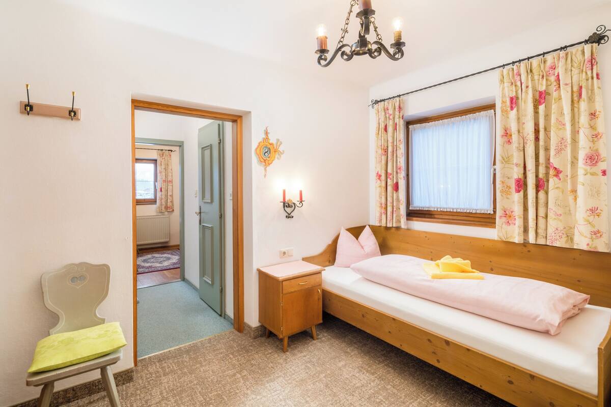 Quarto com cama de madeira, mesa de cabeceira, cadeira, janela com cortinas florais e um lustre.