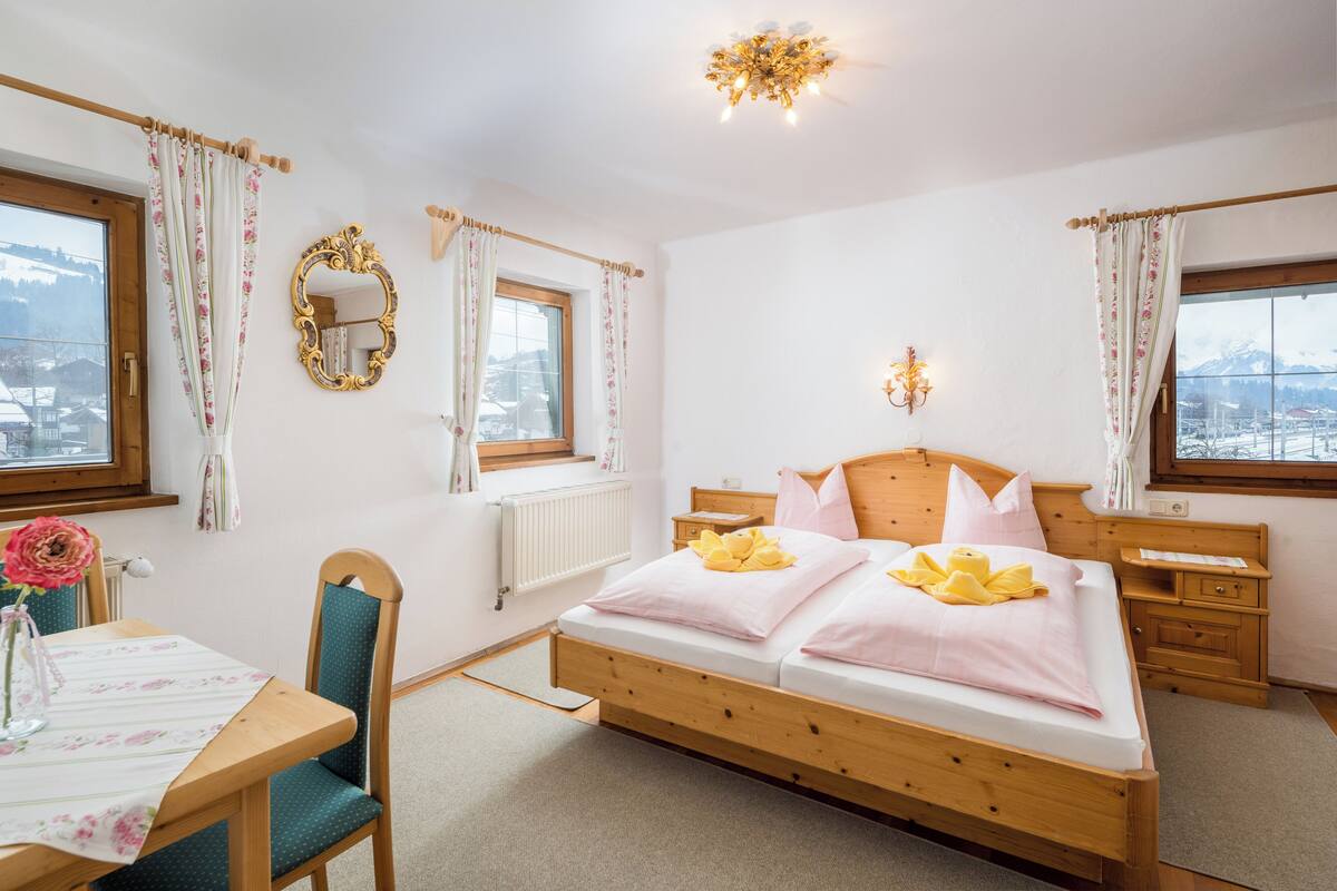 Quarto com cama de madeira, mesa de cabeceira, cadeira, janela com cortinas, espelho e vista exterior.