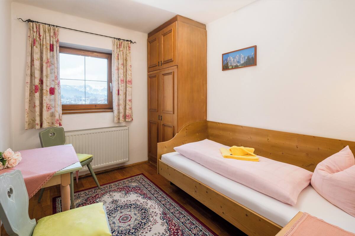 Um quarto pequeno com uma cama de madeira, uma escrivaninha com uma cadeira, um radiador e uma janela com cortinas.