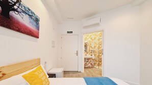 Quarto de hotel com cama, mesa de cabeceira, ar condicionado fixado na parede, porta que dá para um banheiro com cortina de chuveiro florida, e corredor com porta identificada 'IRTEERA'.