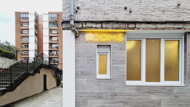 Um edifício com um letreiro 'ROOMS', uma janela com vidro fosco e uma escadaria que leva a uma porta.