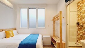 Quarto com cama, um banco, uma mesinha, guarda-roupa e zona de chuveiro.