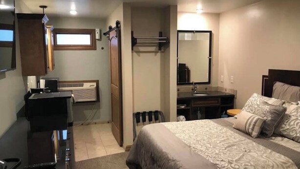 Quarto de hotel com cama, escrivaninha, penteadeira, espelho, janela e banheiro com pia e espelho.