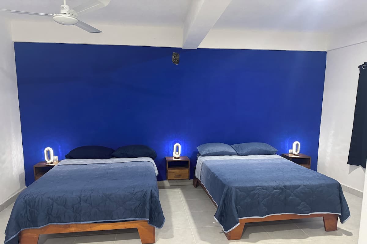 Quarto com duas camas, cada uma com estrutura de madeira e roupa de cama azul. Inclui mesas de cabeceira com arandelas de parede iluminadas.