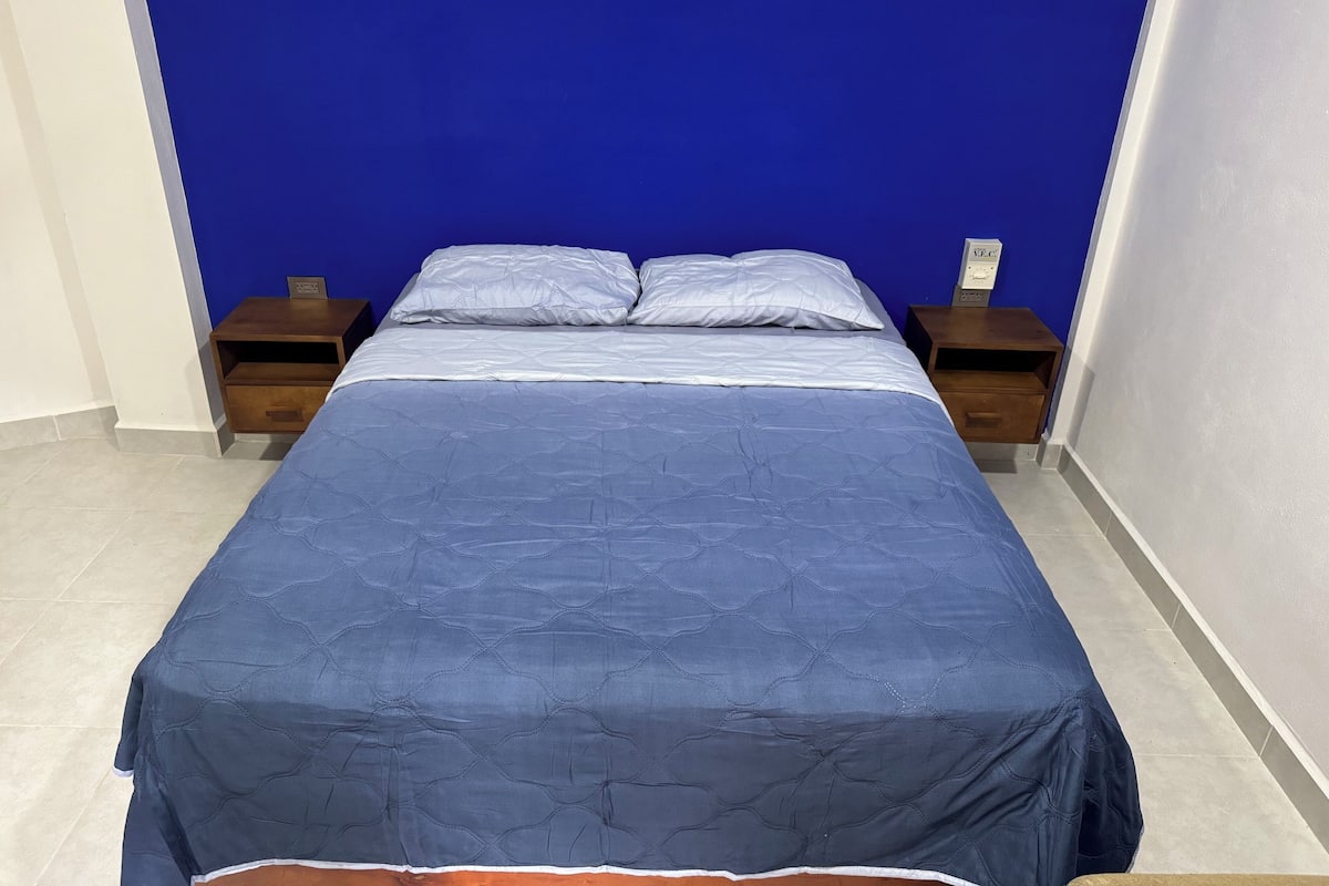 Uma cama com colcha azul, duas mesas de cabeceira de madeira e uma parede azul.