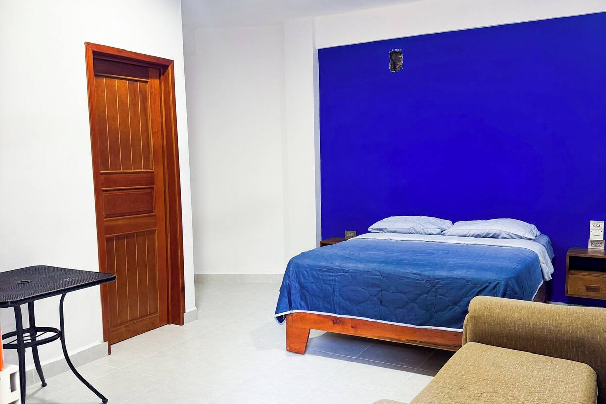 Quarto com cama, porta de madeira, mesa e ventilador de teto.