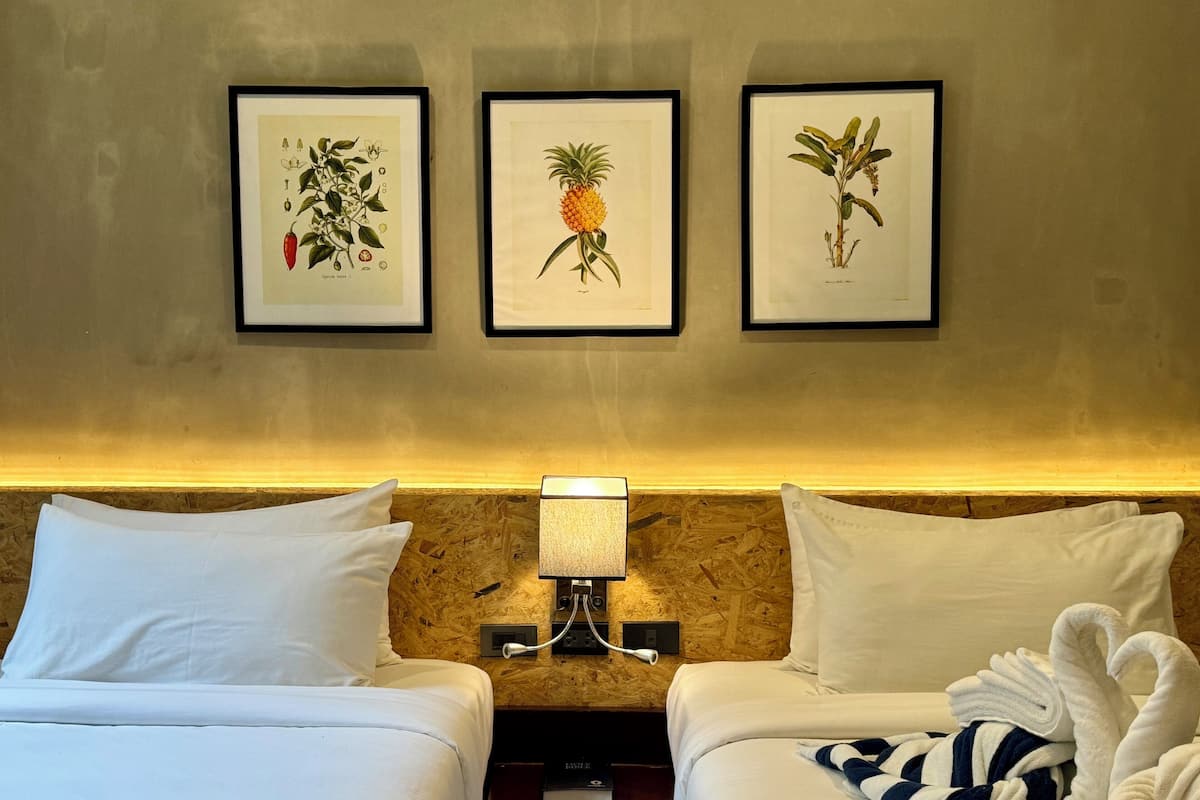 Quarto de hotel com duas camas, uma mesinha de cabeceira, um abajur e três ilustrações botânicas emolduradas na parede.