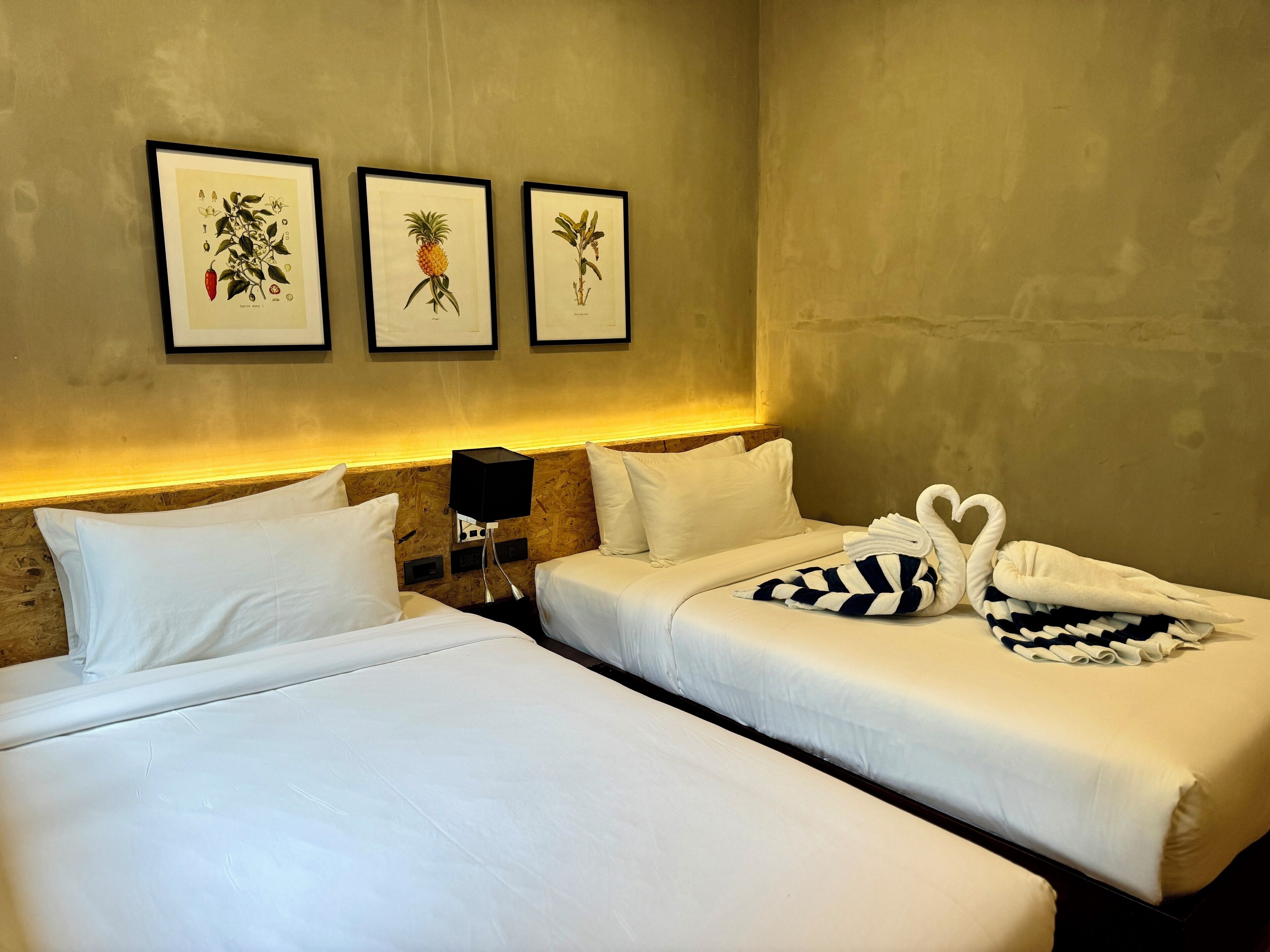 Quarto de hotel com duas camas, roupa de cama branca, luminária fixada na parede e ilustrações botânicas emolduradas.
