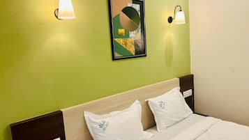 Quarto de hotel com parede verde, pintura abstrata emoldurada, duas luminárias de cabeceira, cama com roupa de cama branca e cabeceira com tomada elétrica.