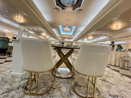 Uma área de refeições luxuosa com piso de mármore, mesas com detalhes dourados e cadeiras brancas.