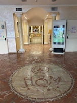 Um lobby com um piso em mosaico circular central, um balcão de recepção e um refrigerador com lista de preços.