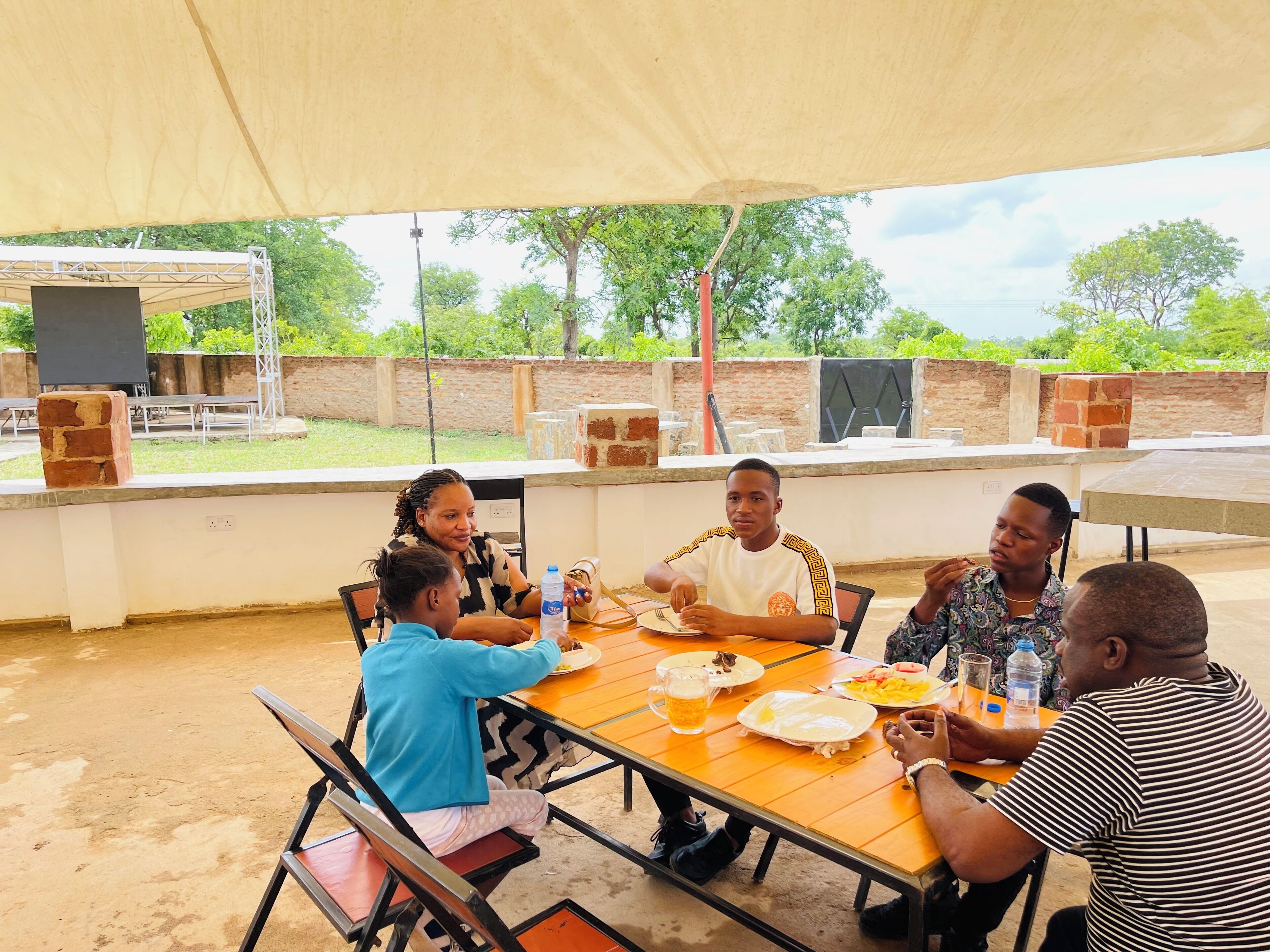 Um grupo de pessoas sentadas em uma mesa ao ar livre, com comida e bebidas, sob um toldo.