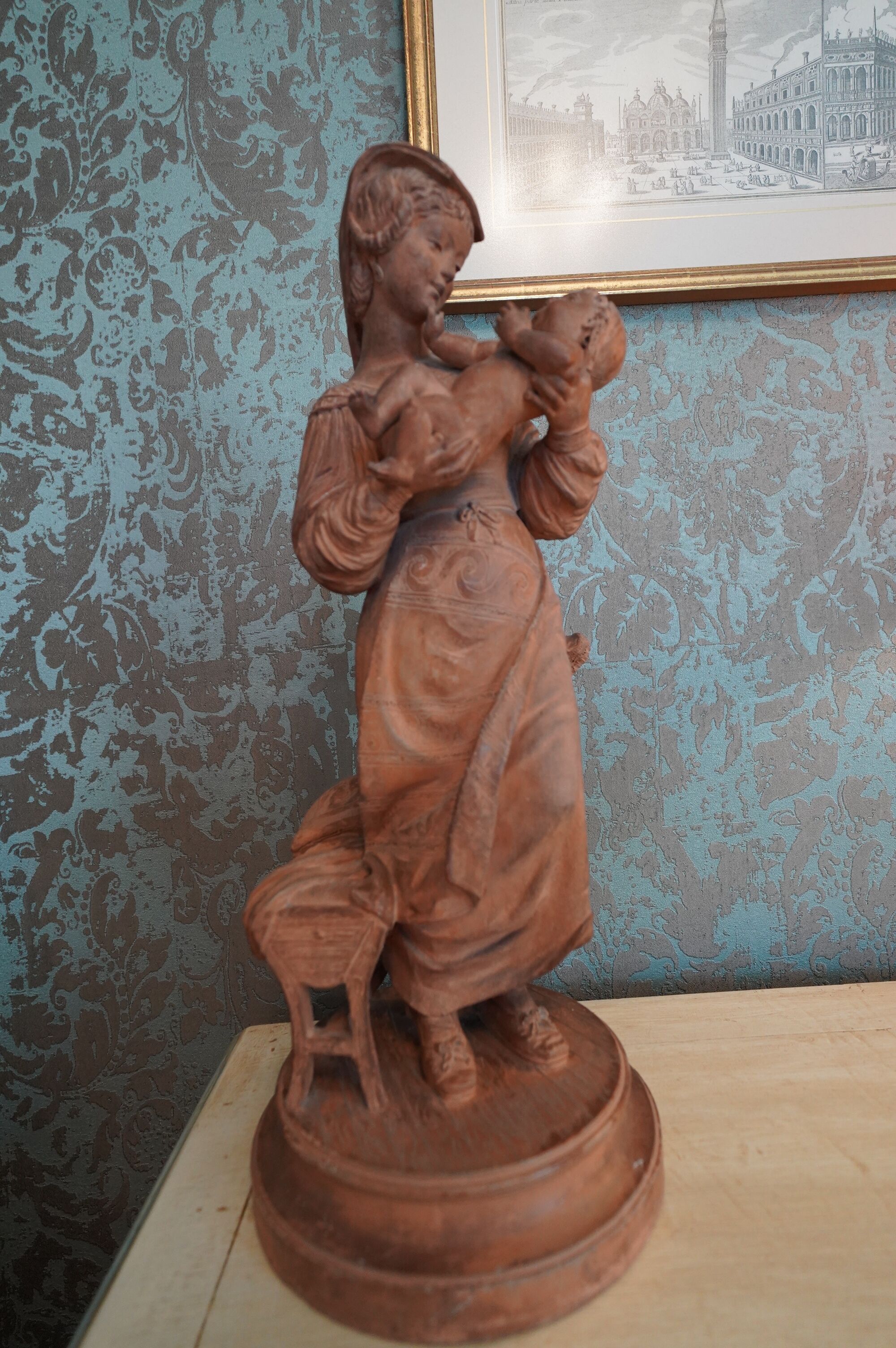 Uma estátua de terracota de uma mulher segurando uma criança, apoiada em uma base de madeira.