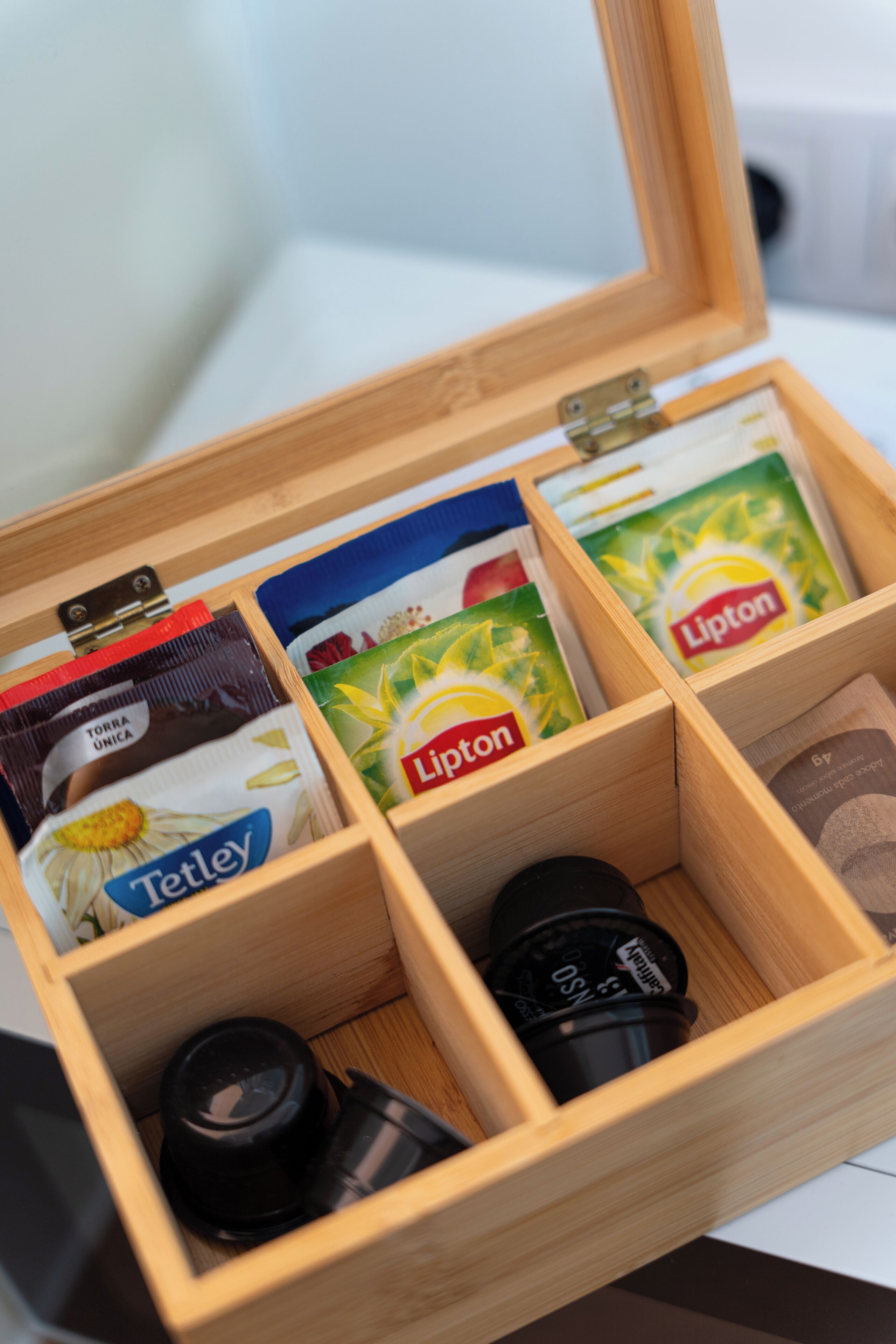 Uma caixa de madeira com divisórias que contêm vários objetos, incluindo saquinhos de chá Lipton e uma lente de câmera.