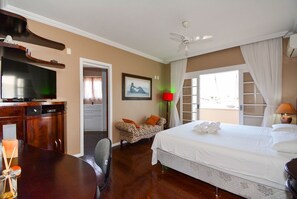 Quarto de hotel com cama, escrivaninha com televisão de tela plana, cadeira, abajur vermelho, quadro na parede e janela com cortinas.