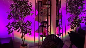 Um quarto com uma poltrona de couro preto, duas plantas em vasos, uma estante com diversos objetos e uma parede com um pôster.
