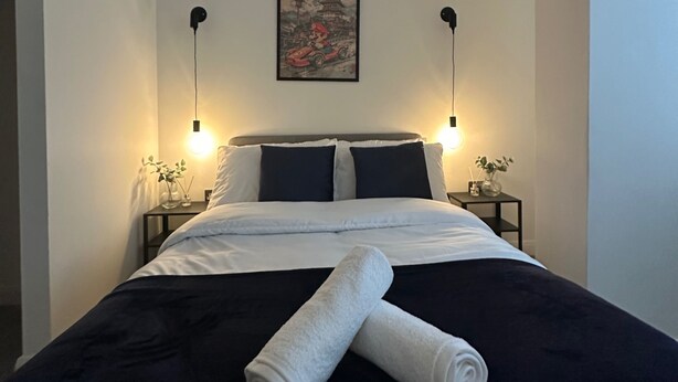 Uma cama bem arrumada com uma toalha enrolada, duas luminárias de cabeceira e um quadro na parede.