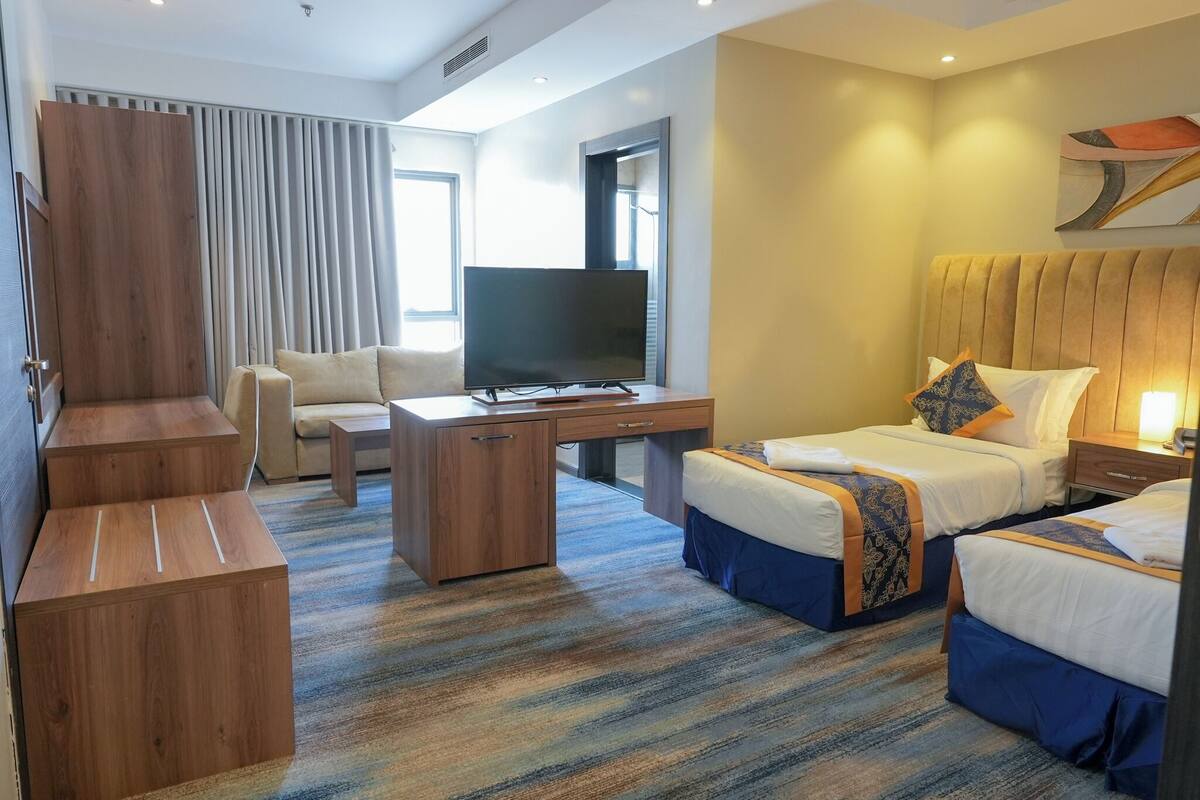 Quarto de hotel com uma cama grande, uma cama menor, uma escrivaninha com televisão, uma cadeira e um guarda-roupa.
