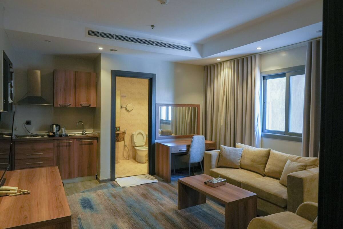 Quarto de hotel com cozinha compacta, sofá, escrivaninha e janela com cortinas.