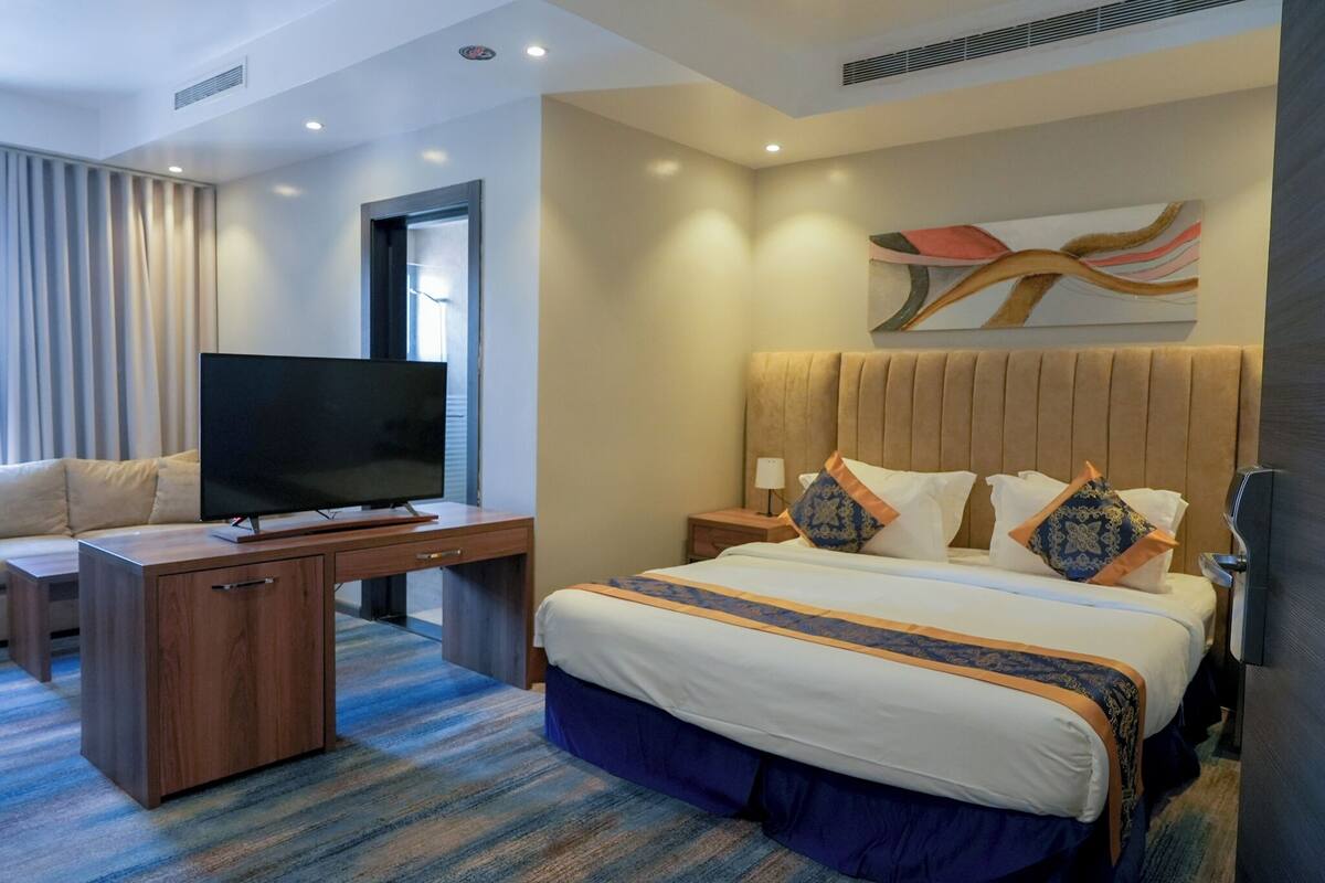 Quarto de hotel com uma cama grande, uma escrivaninha com televisão de tela plana, um sofá e um detalhe decorativo na parede.