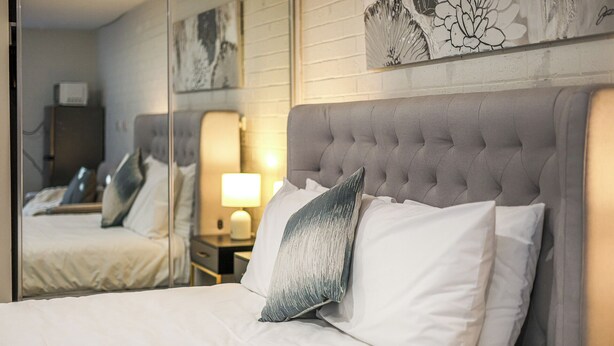 Quarto de hotel com cabeceira estofada, cama com roupa de cama branca e mesa de cabeceira com abajur.