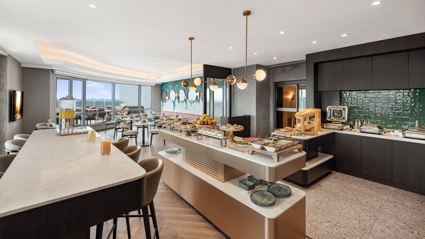 Uma área de jantar moderna em hotel, com uma longa mesa comunitária, um buffet e vista panorâmica da cidade através de grandes janelas.