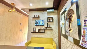 Quarto com sofá amarelo, televisão de tela plana na parede e objetos decorativos nas prateleiras.